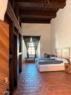 Quarto com piso de tijolos, cama de madeira, mesa de cabeceira e janela com cortinas leves.