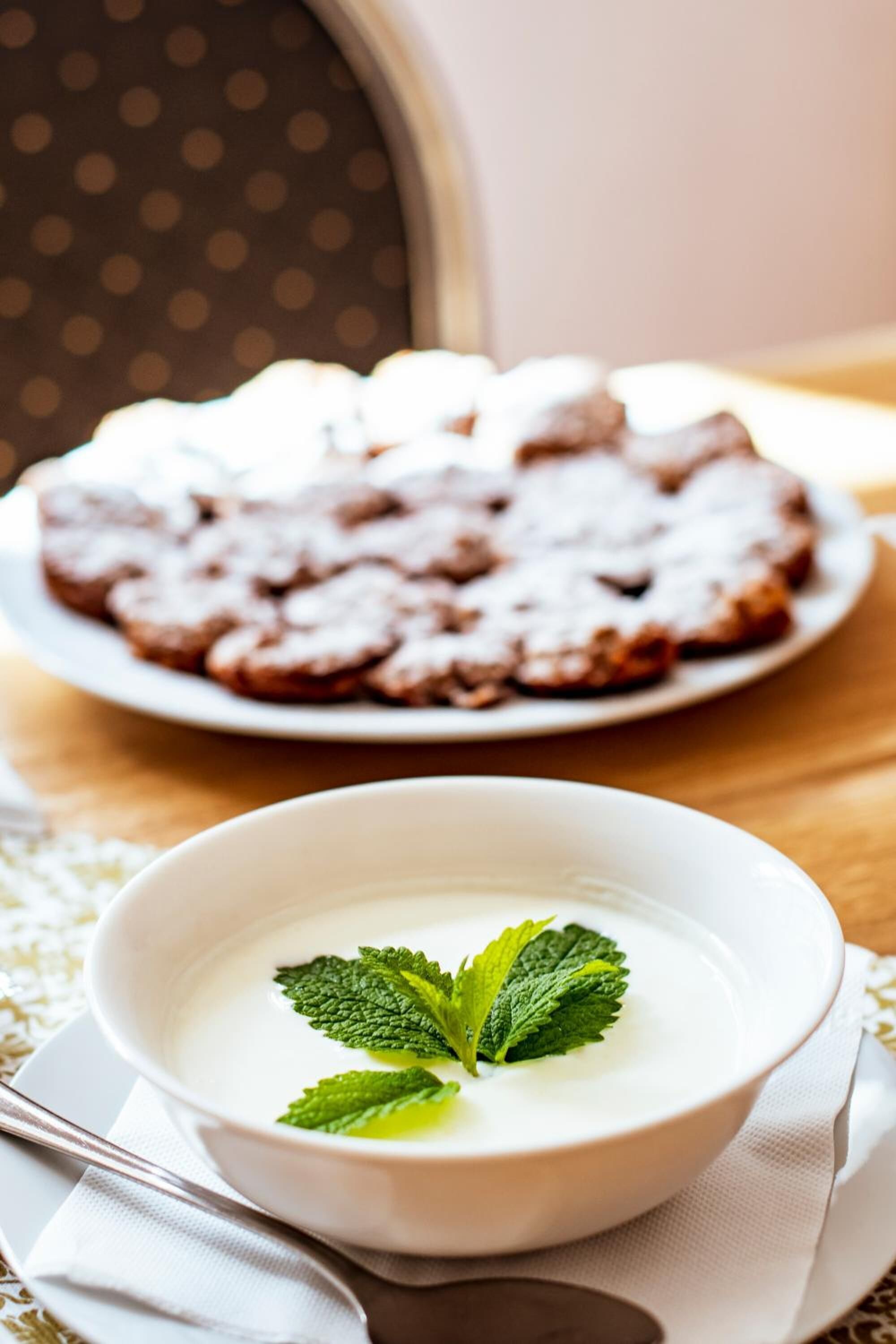 Uma tigela branca de sopa decorada com folhas de hortelã sobre um prato branco, com um prato de biscoitos ao fundo.