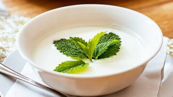 Uma tigela branca de sopa decorada com folhas de hortelã sobre um prato branco, com um prato de biscoitos ao fundo.