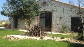 Um edifício rústico de pedra com telhado de telhas, uma grande árvore à frente e um pequeno jardim.