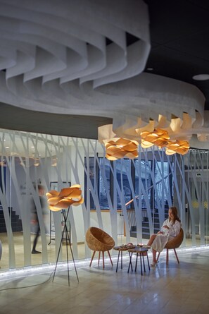 Um interior moderno com uma mulher sentada numa cadeira de vime, uma luminária de chão e arte abstrata nas paredes.