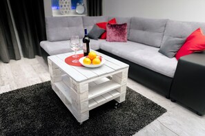 Uma mesa de centro de madeira branca com um jogo americano vermelho, uma garrafa de vinho e uma tigela de frutas numa sala de estar com um sofá cinzento e almofadas vermelhas.