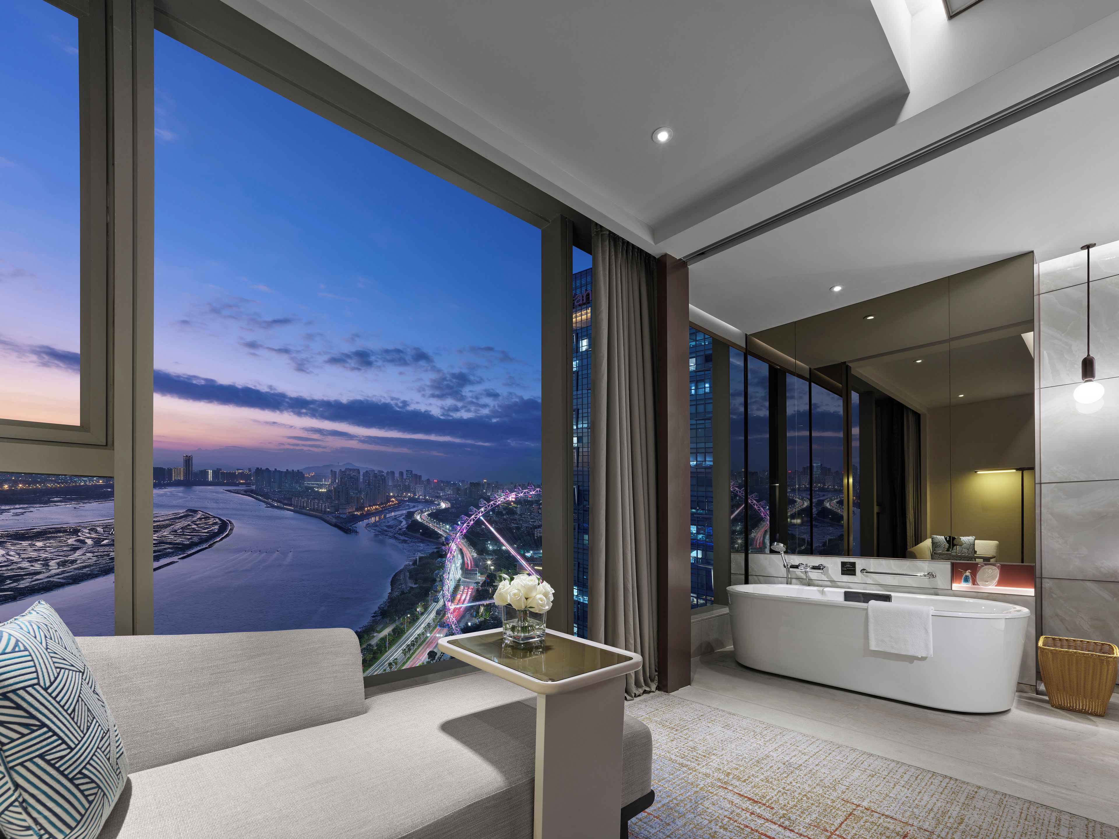 Quarto de hotel moderno com uma janela ampla que oferece vista para a cidade, um sofá, uma mesa pequena com um vaso e um banheiro separado.