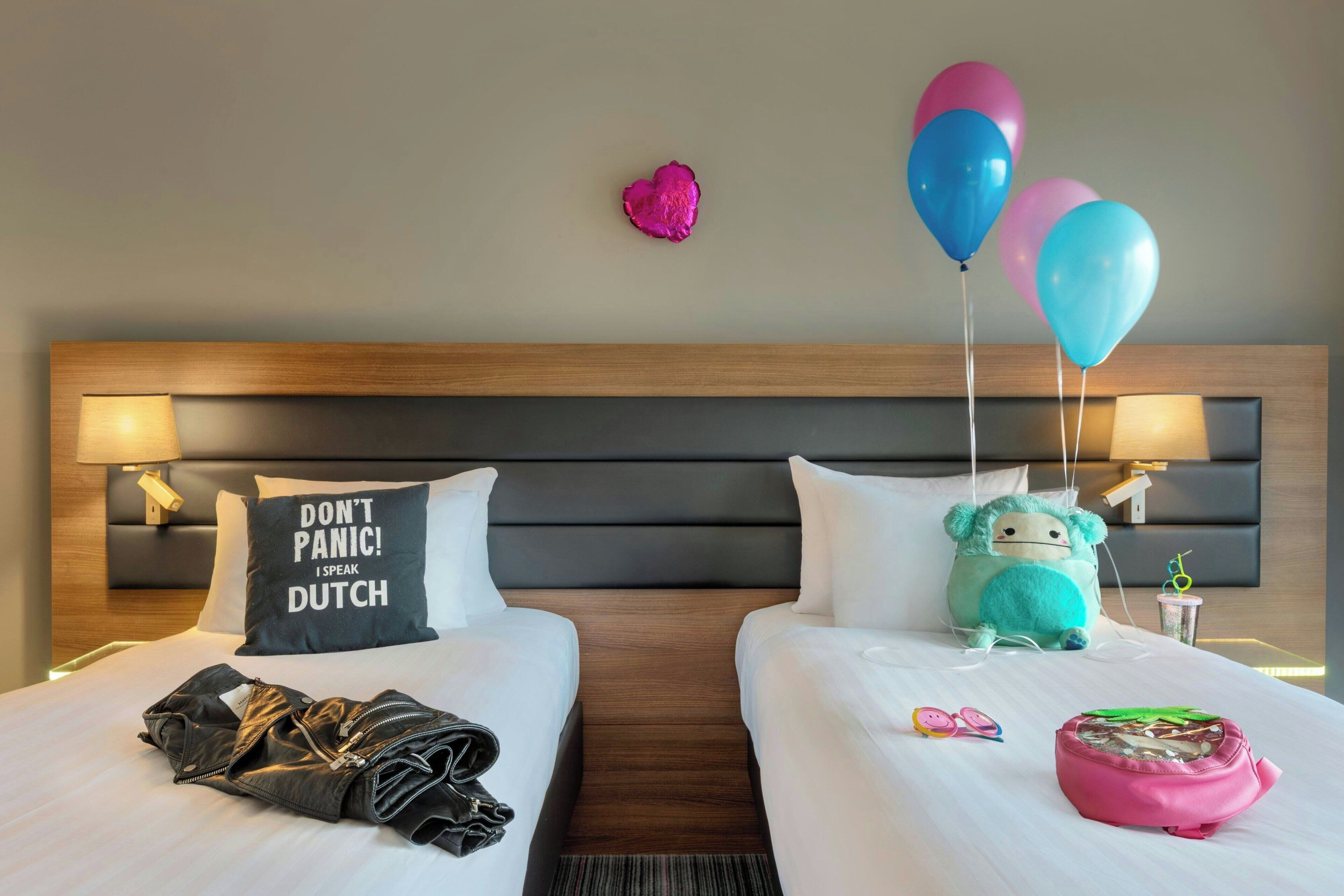 Quarto de hotel com duas camas, cabeceira de madeira, um brinquedo de pelúcia, uma bolsa rosa e balões.