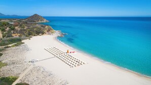Uma praia com fileiras de espreguiçadeiras e guarda-sóis, uma torre salva-vidas e montanhas ao fundo.