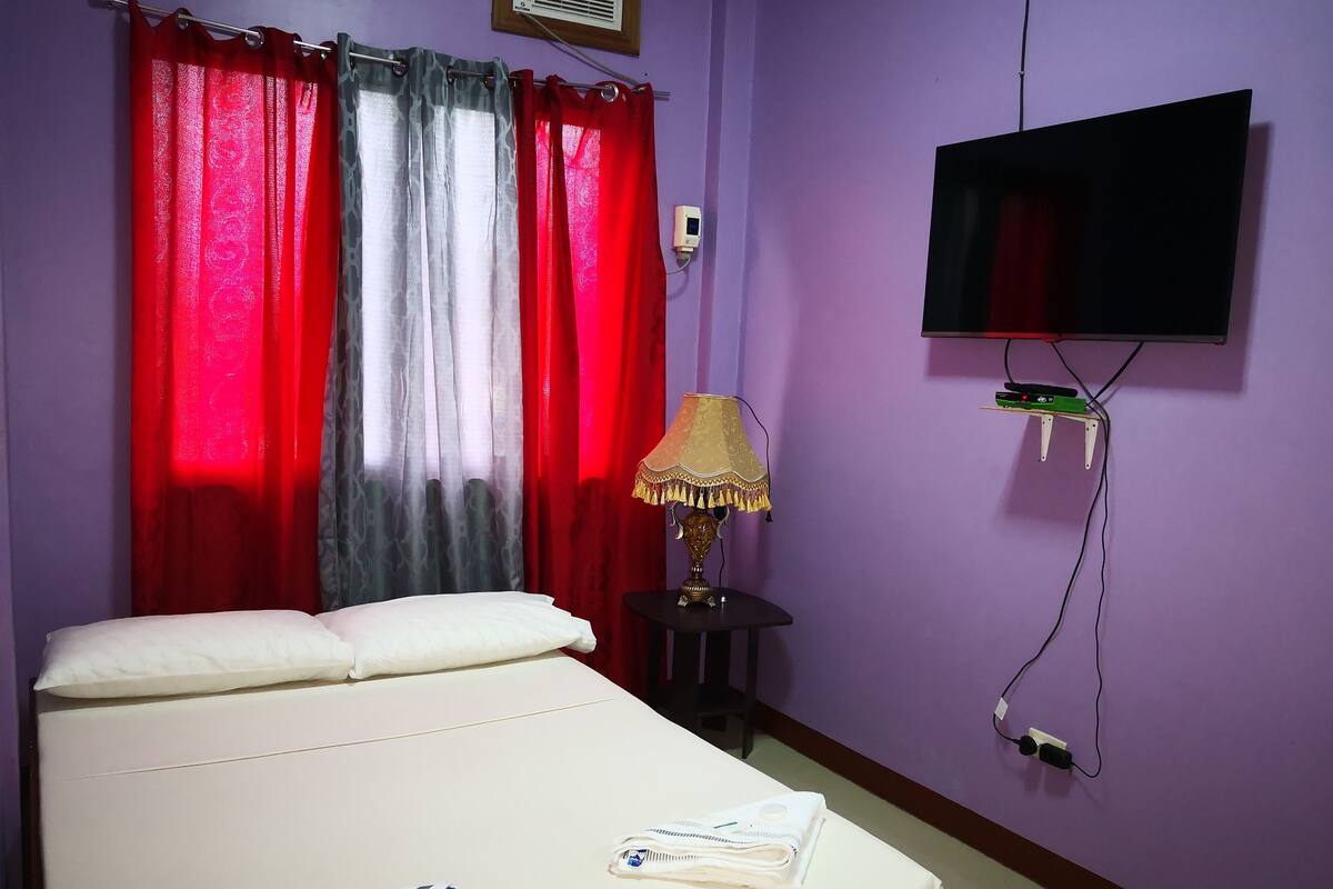 Quarto de hotel com cama, televisão na parede e abajur em uma mesa lateral.
