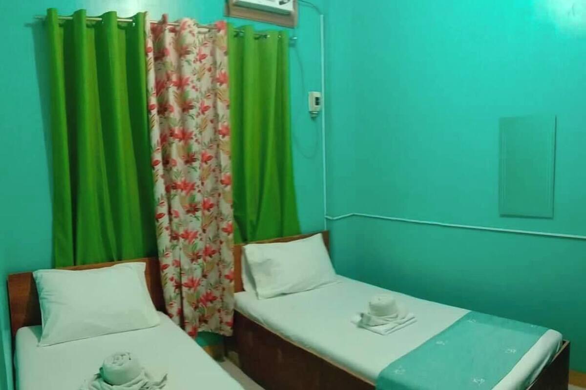 Quarto com duas camas de solteiro, cortinas verdes, cortina com padrão floral e um ar condicionado branco na parede.