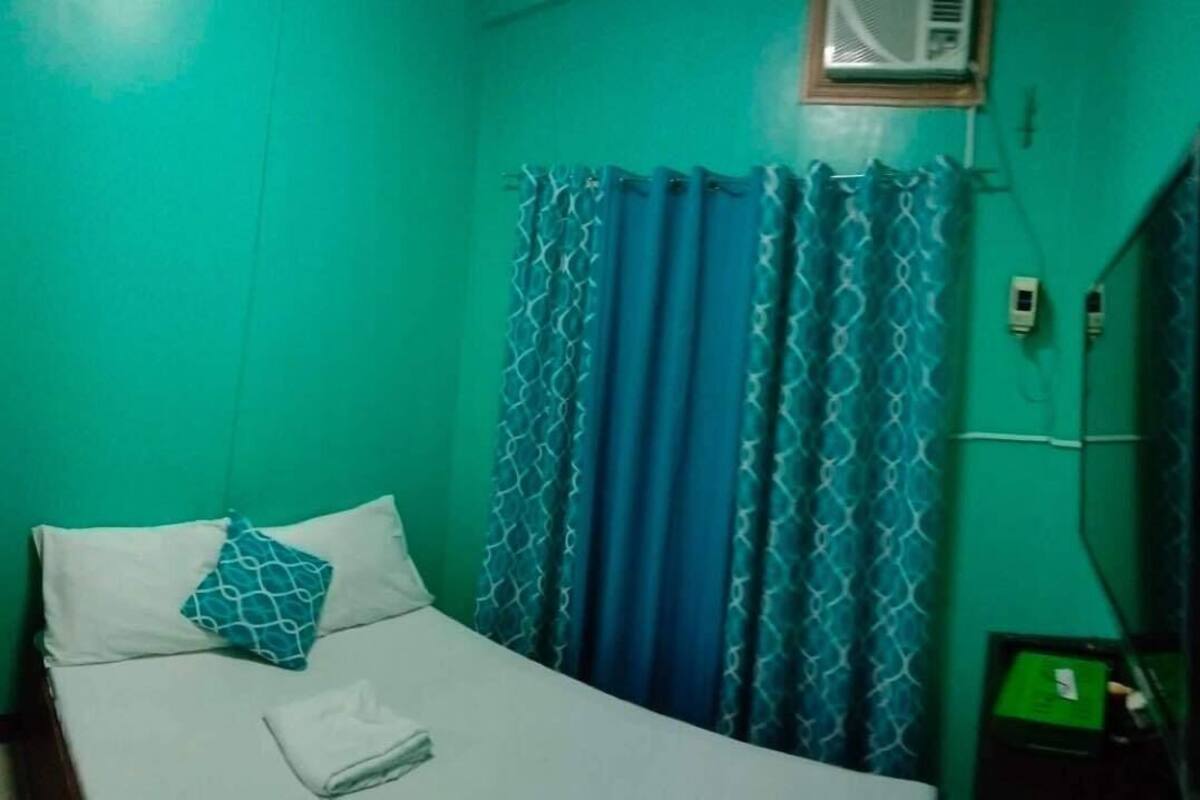 Quarto com paredes verde-água, cama com roupa de cama branca e almofada estampada em azul, janela com cortinas azuis e um pequeno aparelho de ar condicionado acima da janela.
