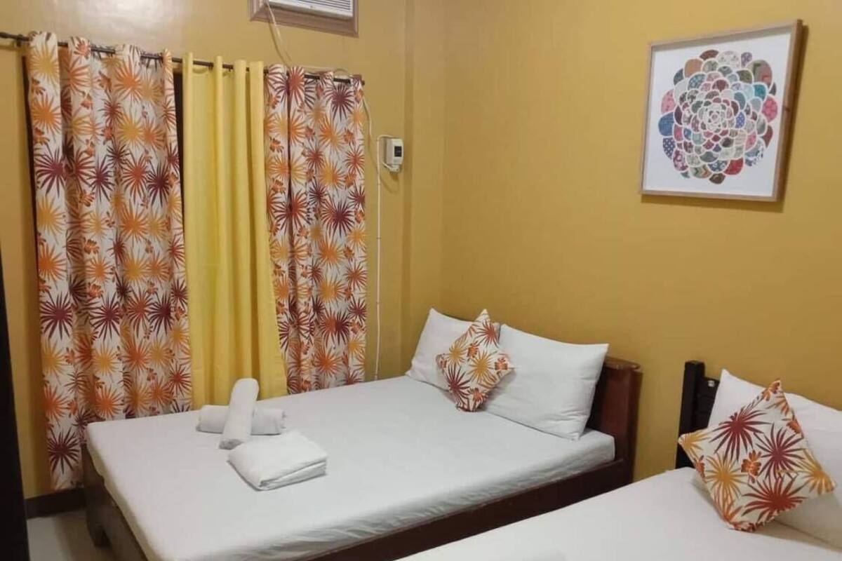 Quarto de hotel com duas camas, cada uma com roupa de cama branca e almofadas combinando. As paredes são amarelas e há uma obra de arte emoldurada na parede.