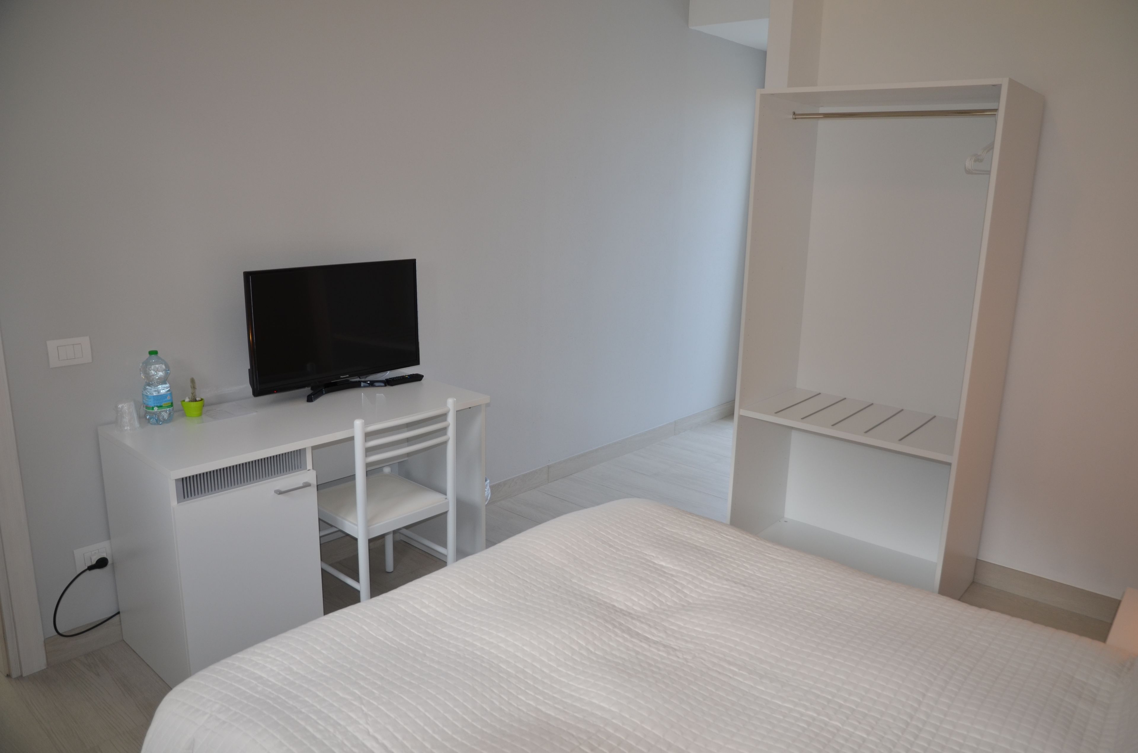 Quarto com cama, televisão sobre uma escrivaninha branca, guarda-roupa e uma garrafa de água.