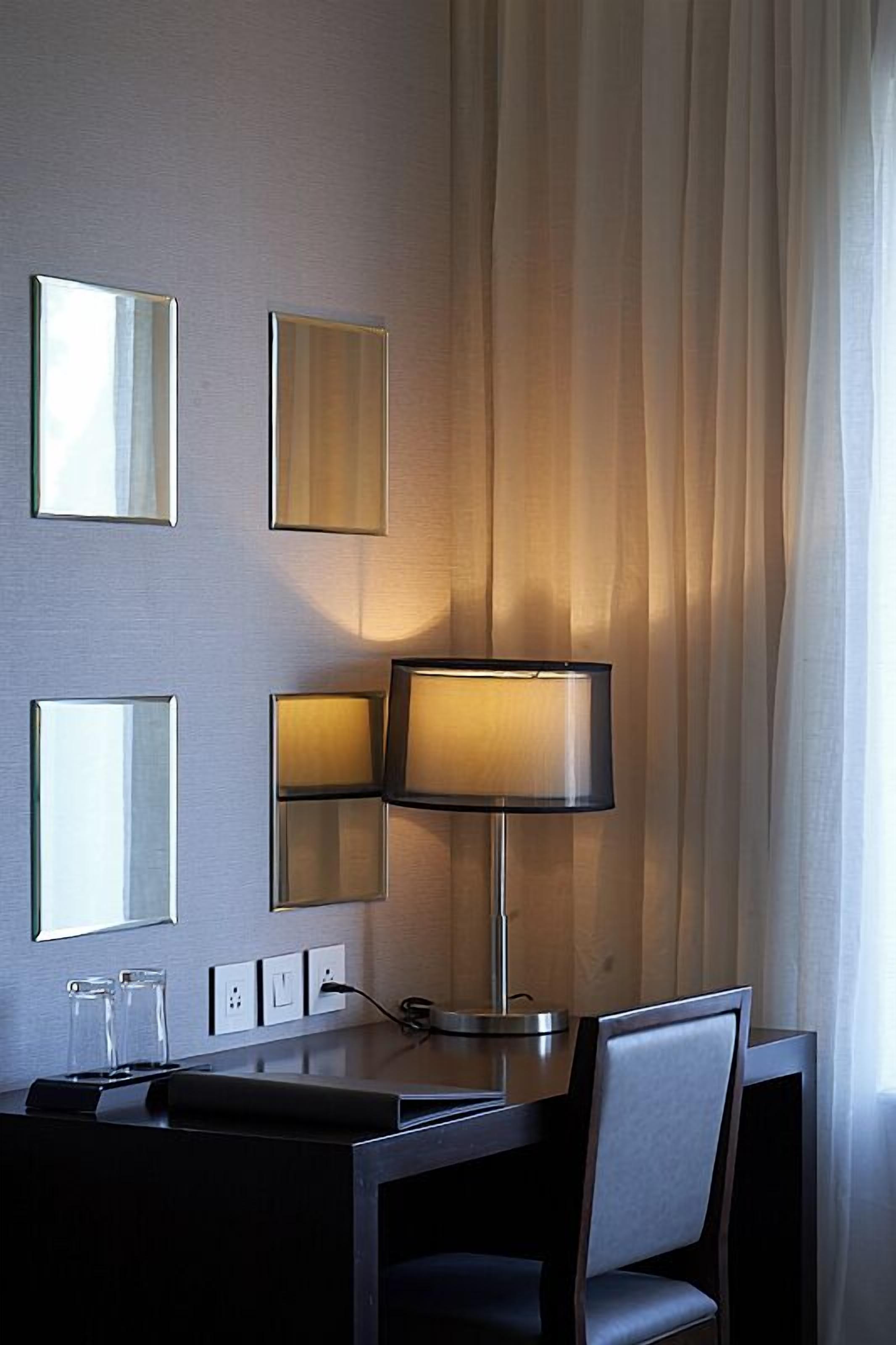 Quarto de hotel com escrivaninha, duas luminárias na parede, um espelho e uma cortina.