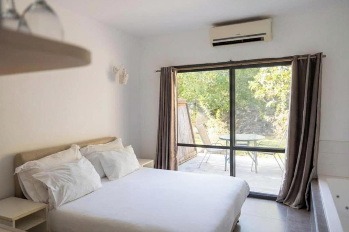 Quarto com cama, mesa de cabeceira, ar condicionado e vista para o exterior através de uma grande janela.
