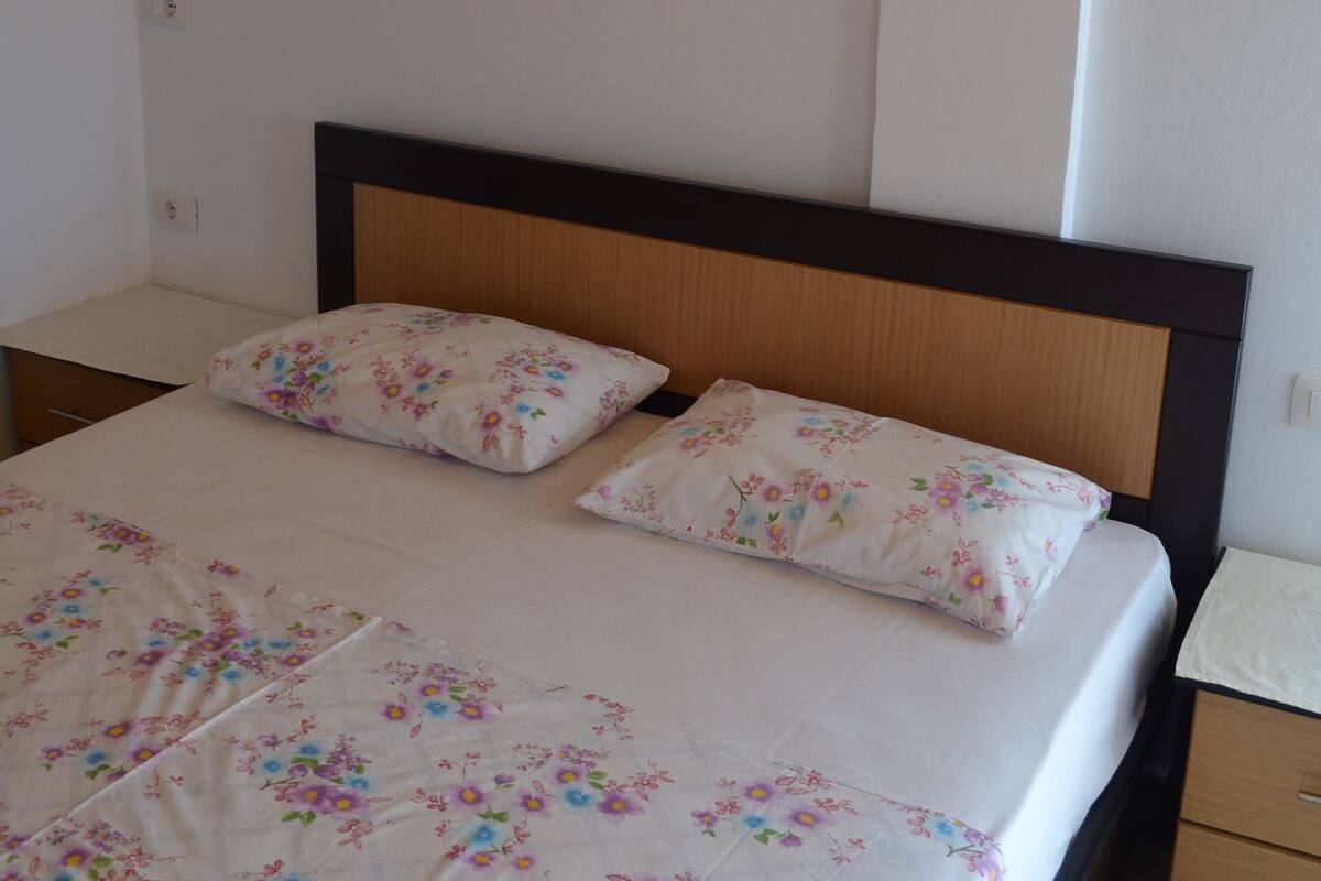 Quarto com cama, colcha floral, cabeceira de madeira, mesa de cabeceira e ar condicionado fixado na parede.