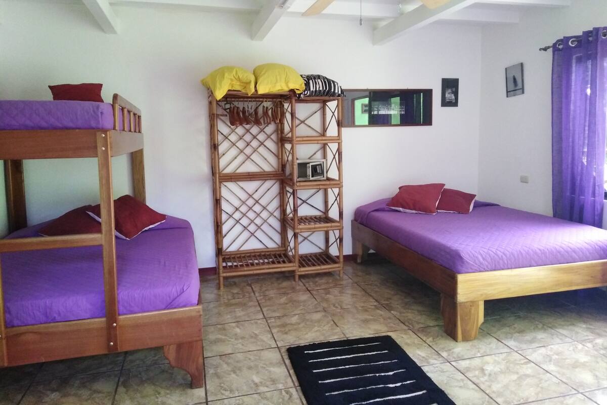 Um dormitório com duas beliches, uma estante de madeira, um ventilador de teto e uma janela com cortinas roxas.