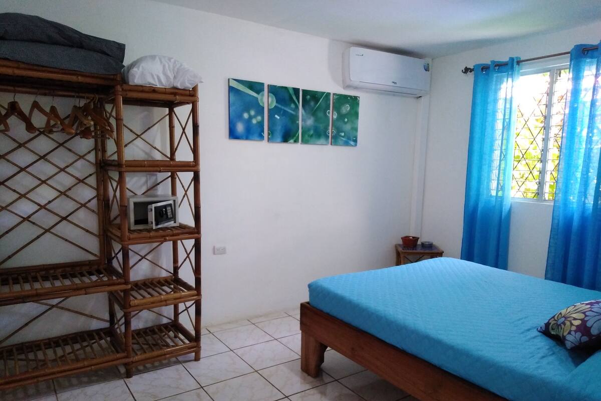 Quarto com cama, prateleira com micro-ondas, janela com cortinas azuis e ar condicionado na parede.