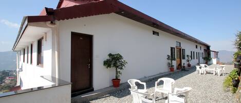 Um edifício com telhado vermelho e paredes brancas, uma porta e várias plantas em vasos. Há cadeiras e mesas de plástico branco do lado de fora.
