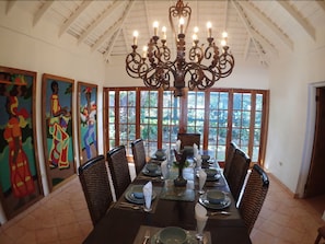 Uma sala de jantar com teto alto e lustre, grandes janelas com vista para o verde, e obras de arte coloridas nas paredes.