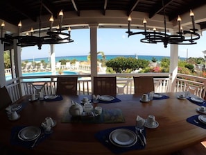 Área de refeições com mesa posta para quatro pessoas, com vista para uma piscina e para o oceano.