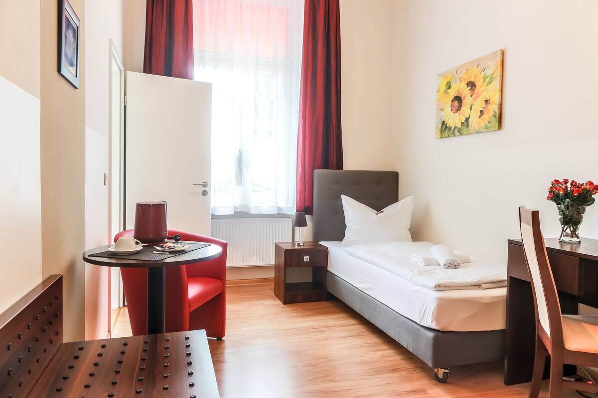 Quarto de hotel pequeno com uma cama, uma mesinha com uma cadeira, uma mesinha de cabeceira com abajur e uma janela com cortinas.