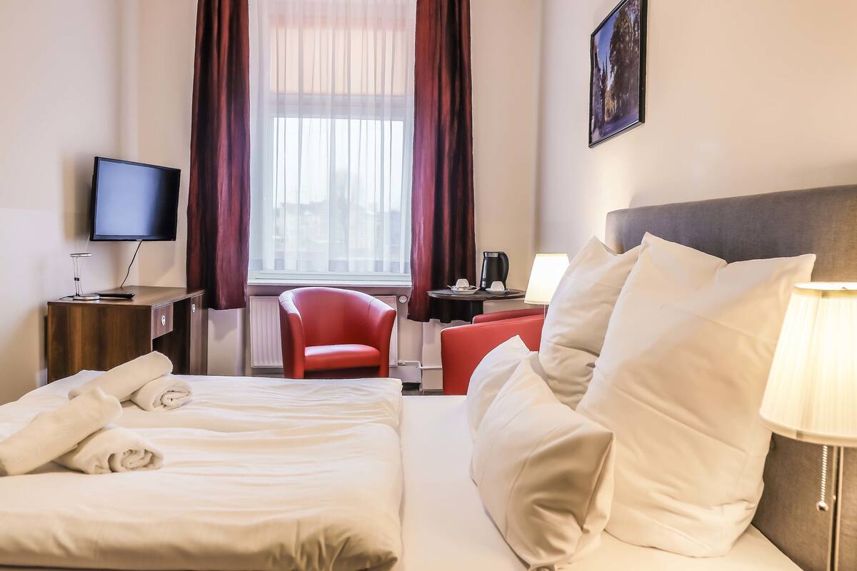Quarto de hotel com cama, escrivaninha com televisão, cadeira, abajur e janela com cortinas.