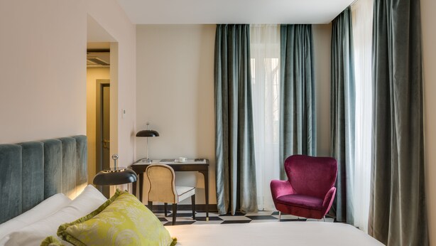 Quarto de hotel com cama, escrivaninha, cadeira e janela com cortinas.