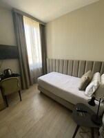 Quarto de hotel com cama, escrivaninha, cadeira, televisão e janela com cortinas.