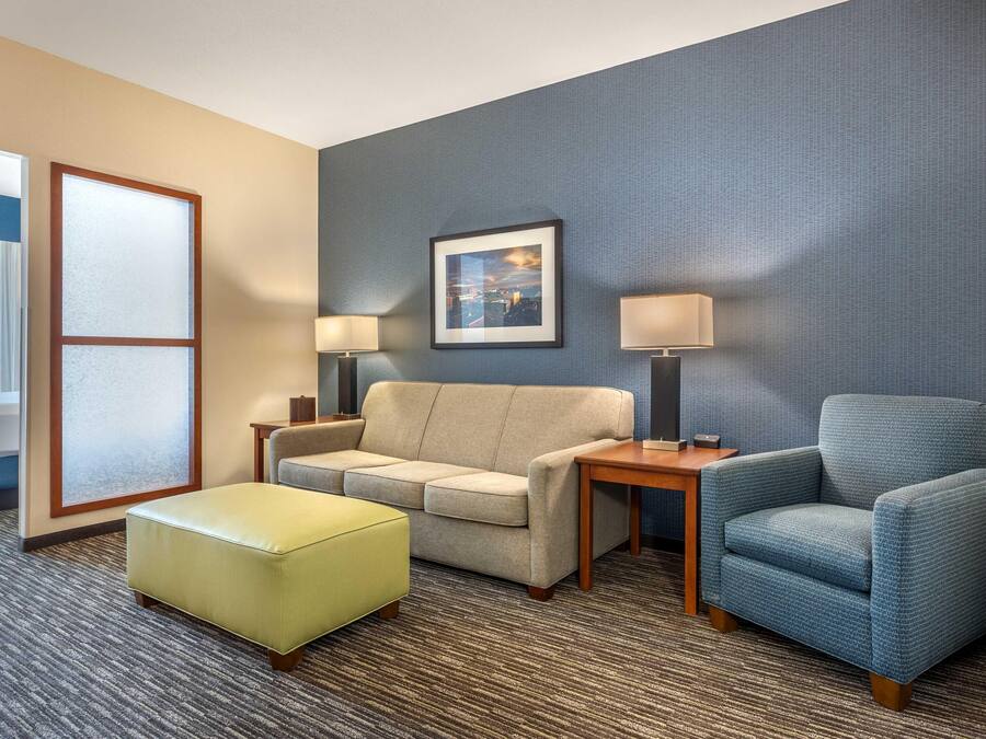 Quarto de hotel com um sofá bege, uma poltrona azul, um banquinho amarelo, duas mesas laterais com luminárias e um quadro na parede.