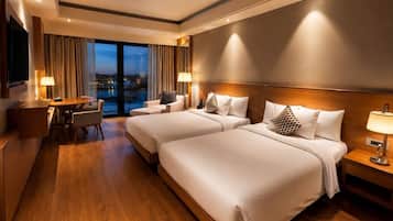 Quarto de hotel com duas camas, uma escrivaninha e vista para a cidade ao entardecer.
