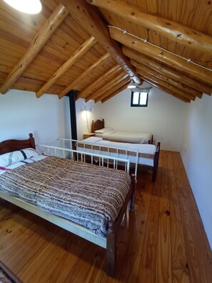 Quarto com piso de madeira, teto inclinado, duas camas, uma janela pequena e um abajur fixado na parede.