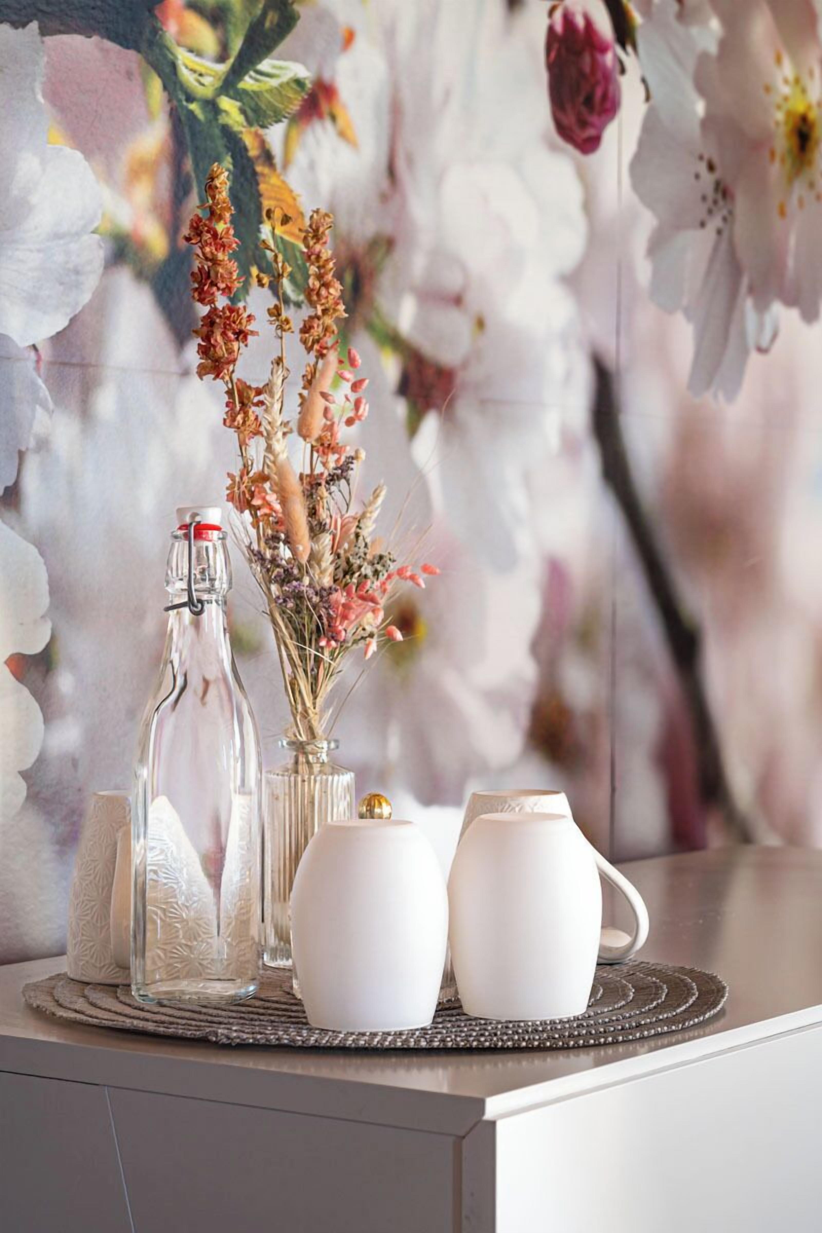 Uma mesa com uma garrafa de vidro transparente, um vaso branco com flores secas e um jarro branco sobre um jogo americano trançado.