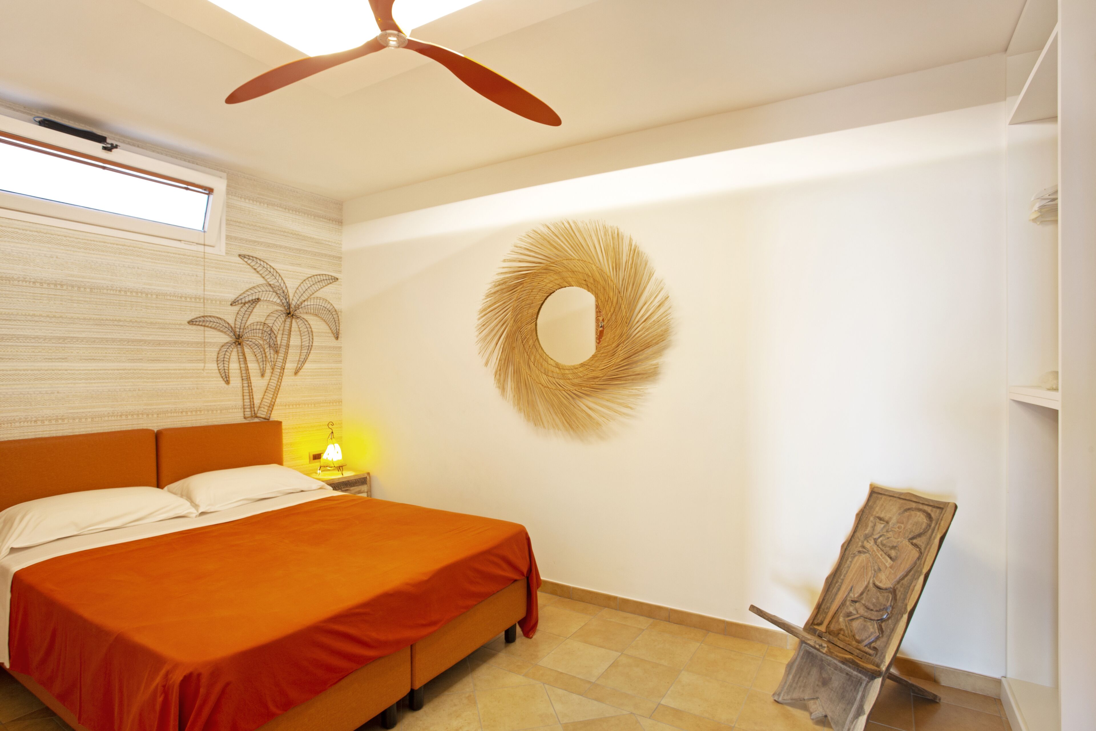 Quarto com cama, ventoinha no teto, espelho fixado na parede e uma mesinha de cabeceira com abajur.