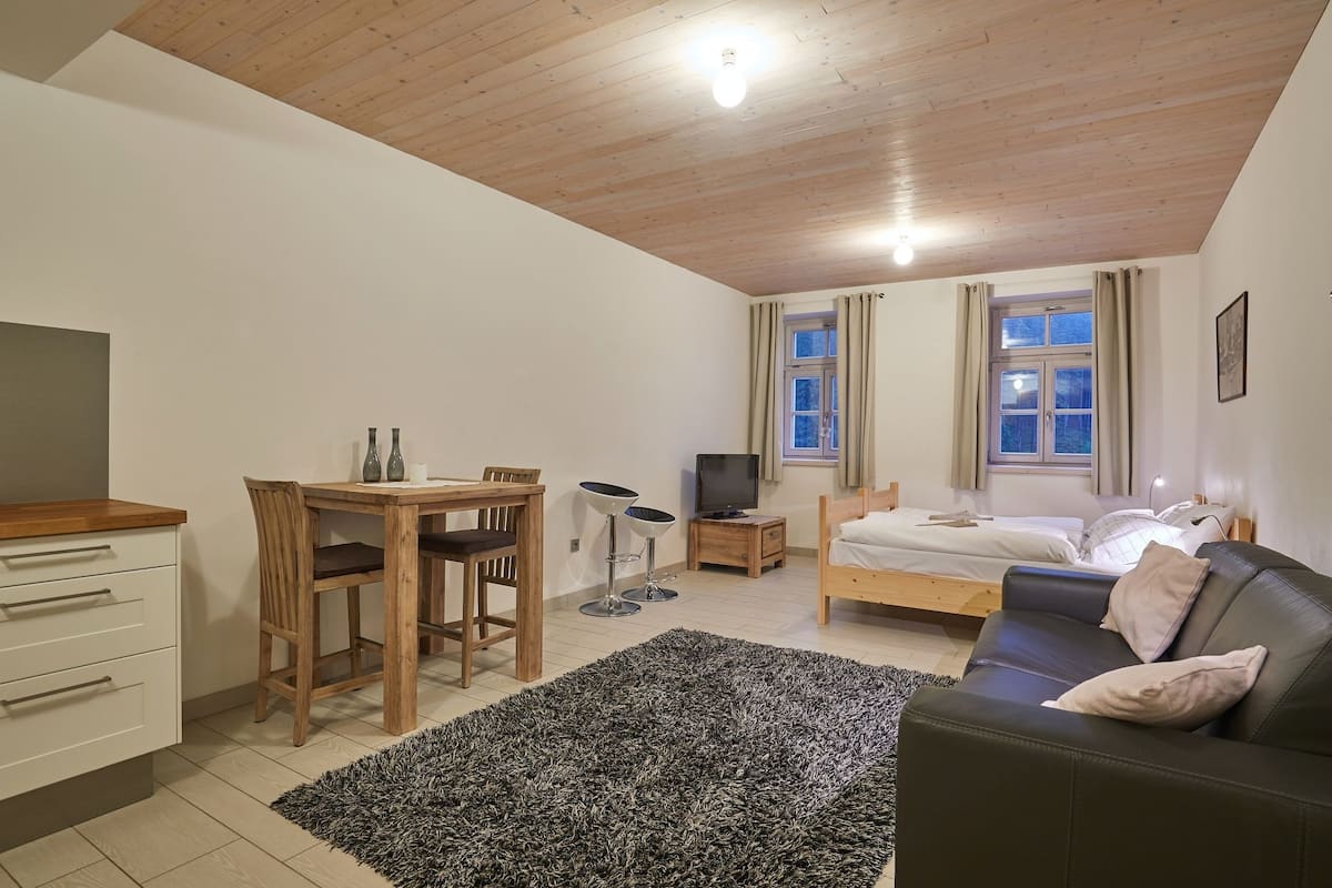 Quarto com teto de madeira, cama com roupa de cama branca, sofá de couro preto, mesa de jantar com cadeiras, TV sobre um suporte e janela com cortinas.