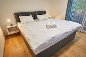 Cama bem arrumada com lençóis brancos e duas toalhas enroladas, mesa de cabeceira com abajur e vista para uma varanda com cortinas azuis.
