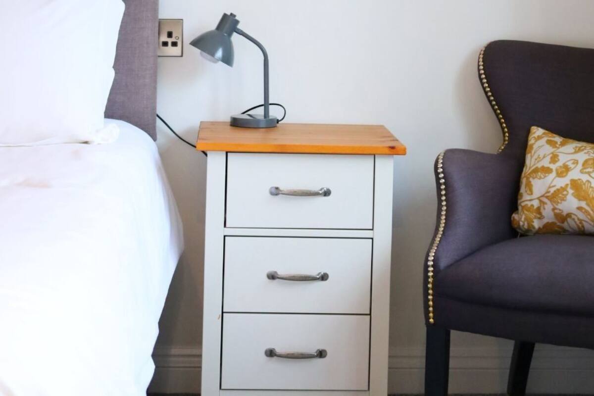 Quarto de hotel com cama, mesa de cabeceira com luminária, uma cadeira e um pequeno armário de madeira com gavetas.