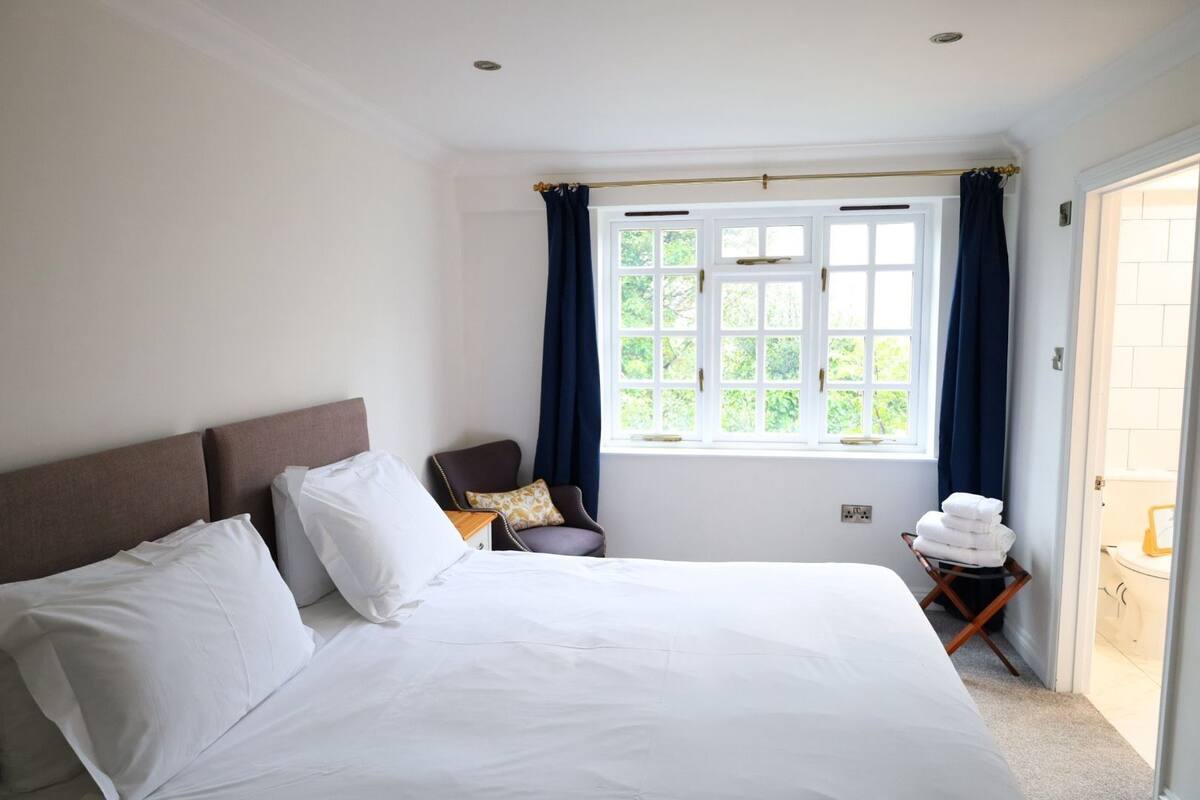 Quarto de hotel com uma cama grande, uma cadeira, uma janela com cortinas e vista para o verde lá fora.
