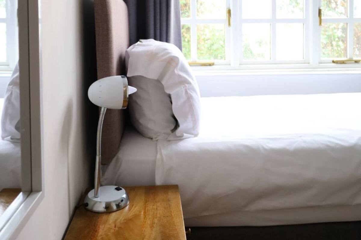 Cama bem arrumada com lençóis brancos, abajur em mesa de cabeceira de madeira e janela com cortinas.
