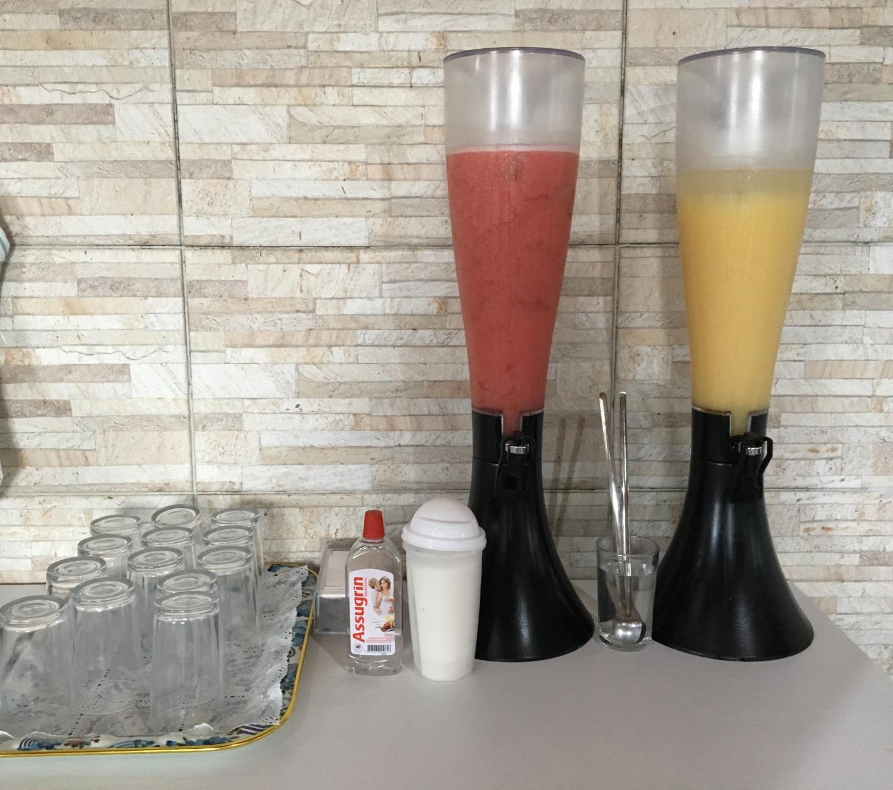 Dois dosadores de bebidas com líquidos de cores diferentes, uma bandeja de copos e uma garrafa de Assugrin.
