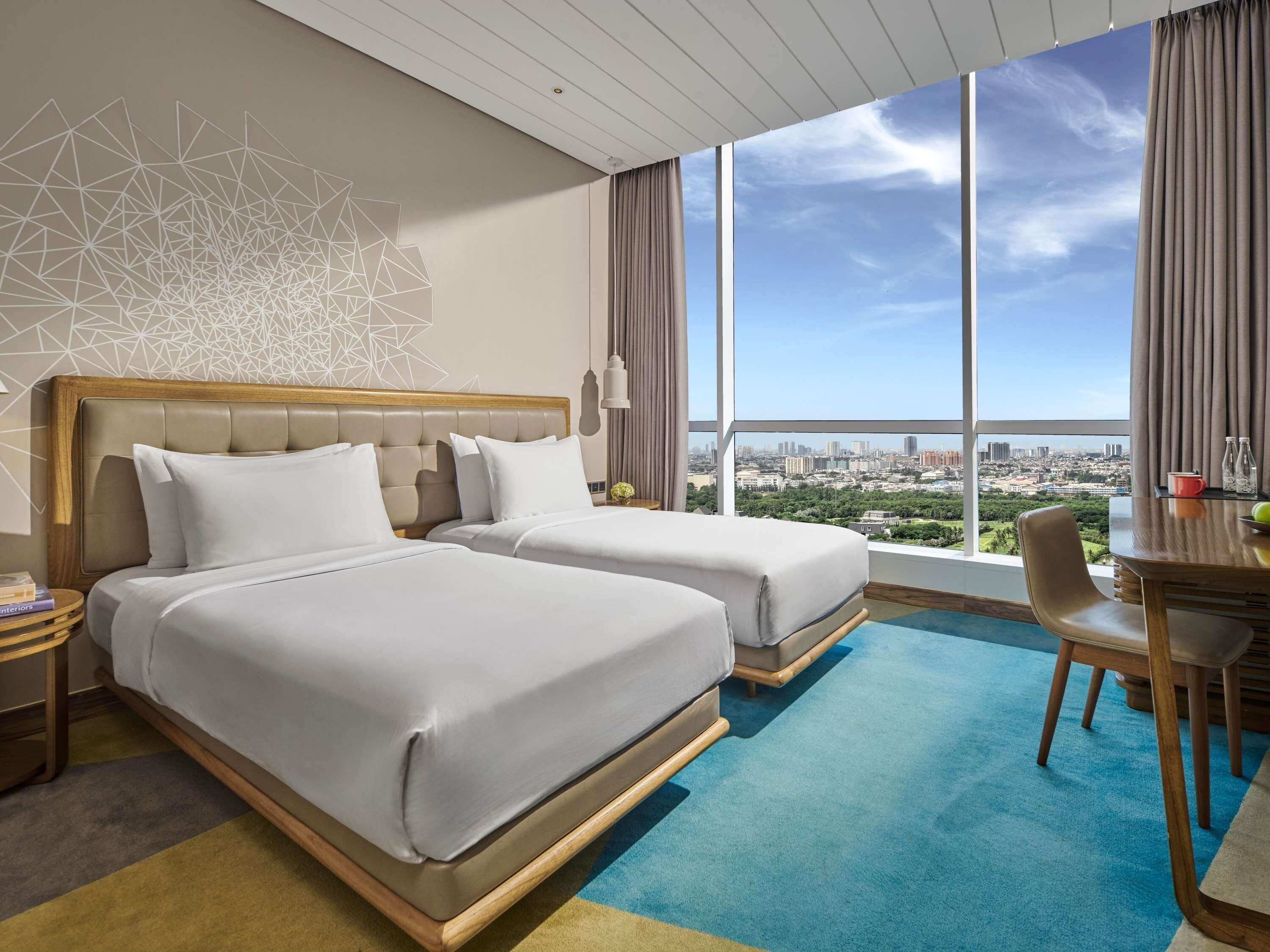 Quarto de hotel com duas camas, uma mesa de jantar, uma cadeira e vista para a cidade, além de um design geométrico na parede.