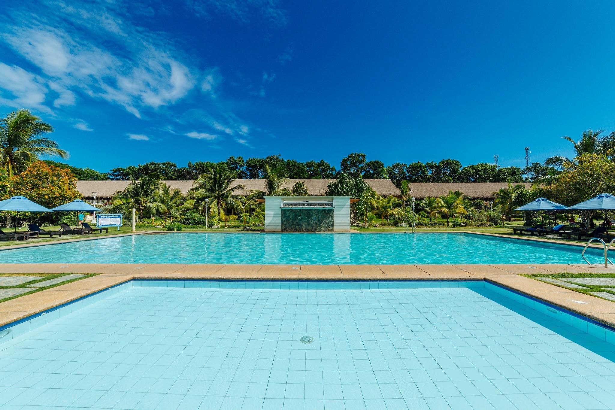 Piscina com bordas azulejadas, rodeada por cadeiras de espreguiçadeira e guarda-sóis, com uma estrutura de telhado de palha ao fundo.