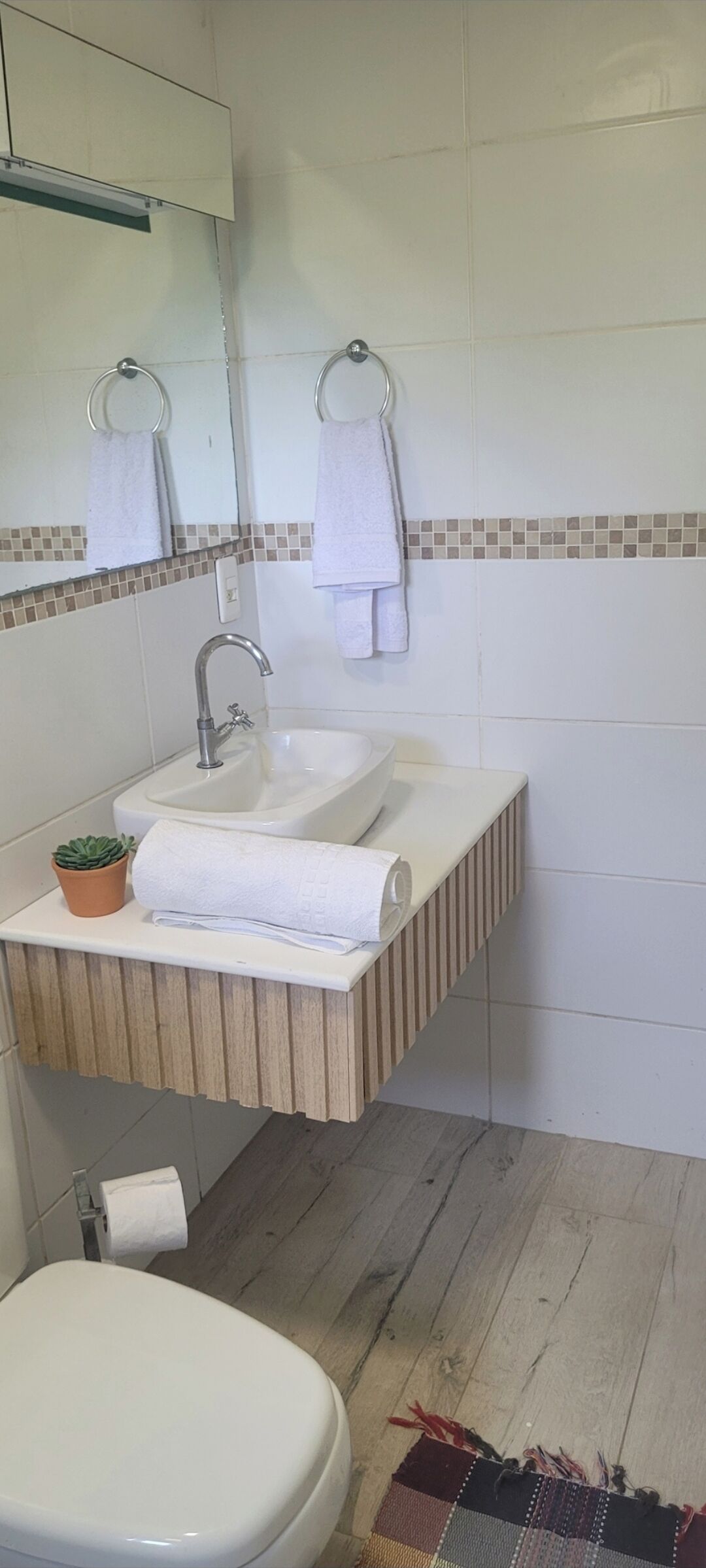 Casa de banho com lavatório branco, espelho, sanita e uma pequena planta em vaso.