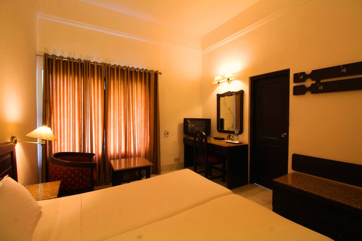 Quarto de hotel com cama, escrivaninha, cadeira, televisão, espelho e porta.