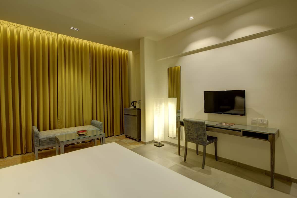 Quarto de hotel com cama, escrivaninha com cadeira, televisão, mesinha de cabeceira com abajur e um sofá.