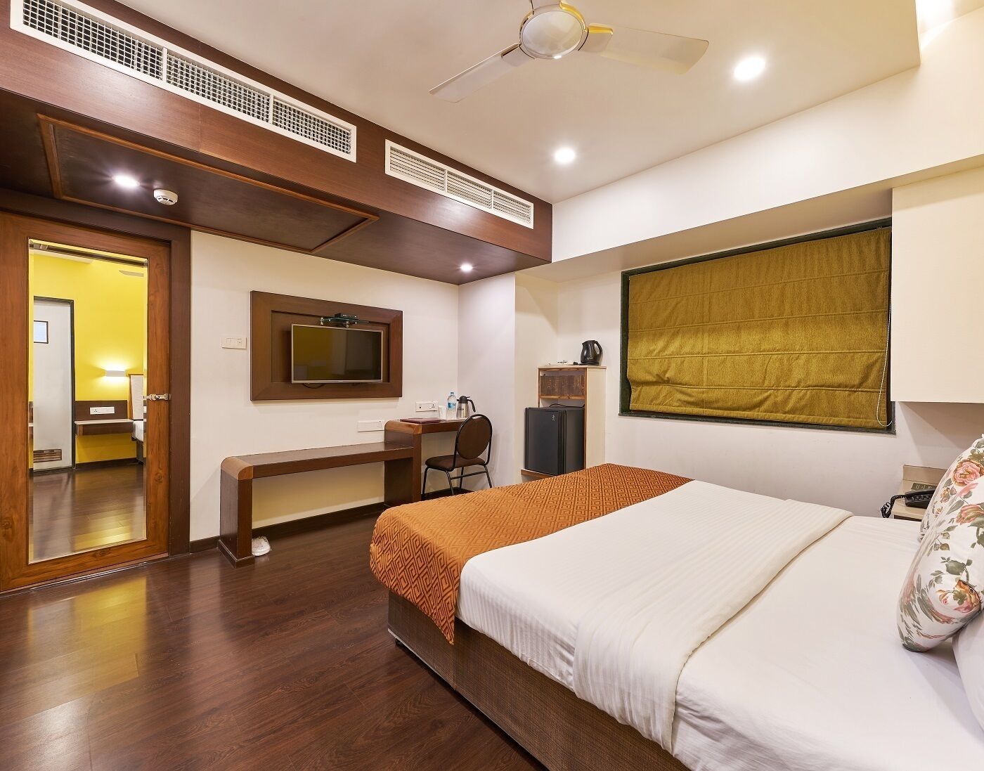 Quarto de hotel com cama, escrivaninha, cadeira, televisão e janela com persiana.