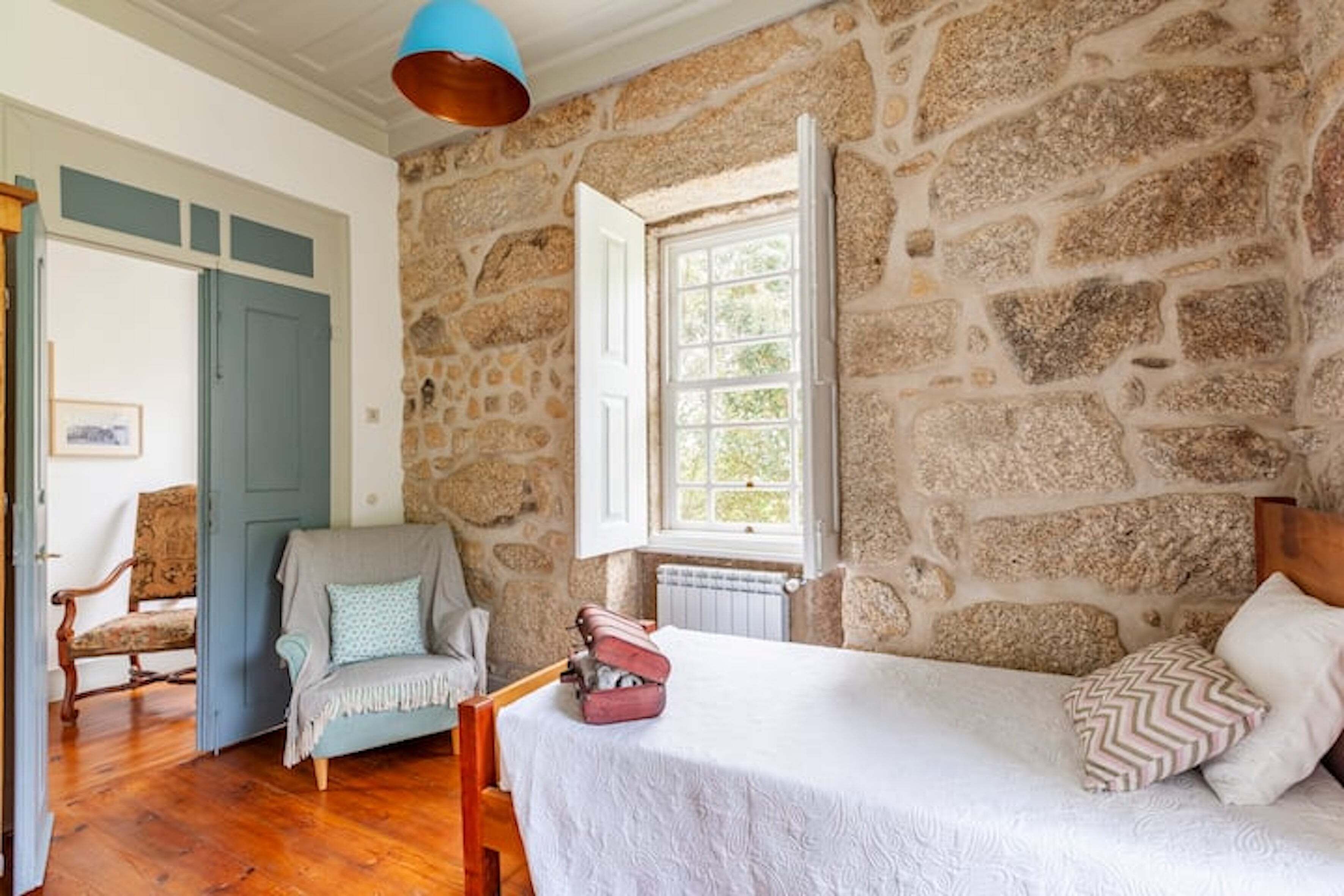 Quarto com parede de pedra, cama com colcha branca, uma cadeira e janela com persianas abertas.
