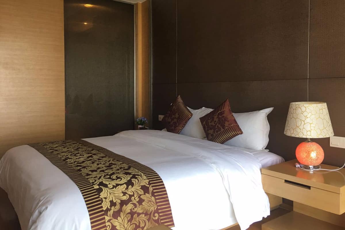 Quarto de hotel com uma cama grande, mesa de cabeceira e abajur, e cabeceira escura.