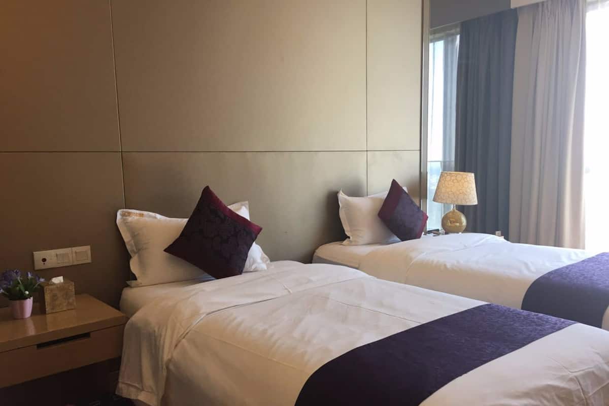 Quarto de hotel com duas camas, uma mesinha de cabeceira com abajur, uma pequena mesa com planta e um par de chinelos no chão.