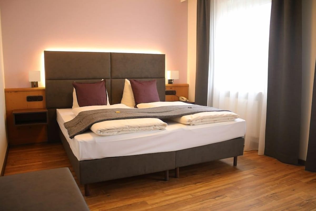Uma cama bem arrumada com almofadas, uma mesa de cabeceira com abajur e piso de madeira.