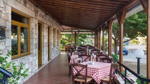 Uma área de refeições ao ar livre coberta, com paredes de pedra, vigas de madeira e toalhas de mesa xadrez.