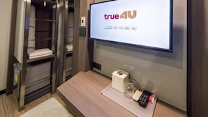 Quarto de hotel com televisão exibindo 'true4U' e uma pequena mesa com controle remoto, óculos e caixa de lenços.