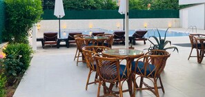 Área de piscina com mobiliário de vime, uma mesa de vidro e um viveiro verde.